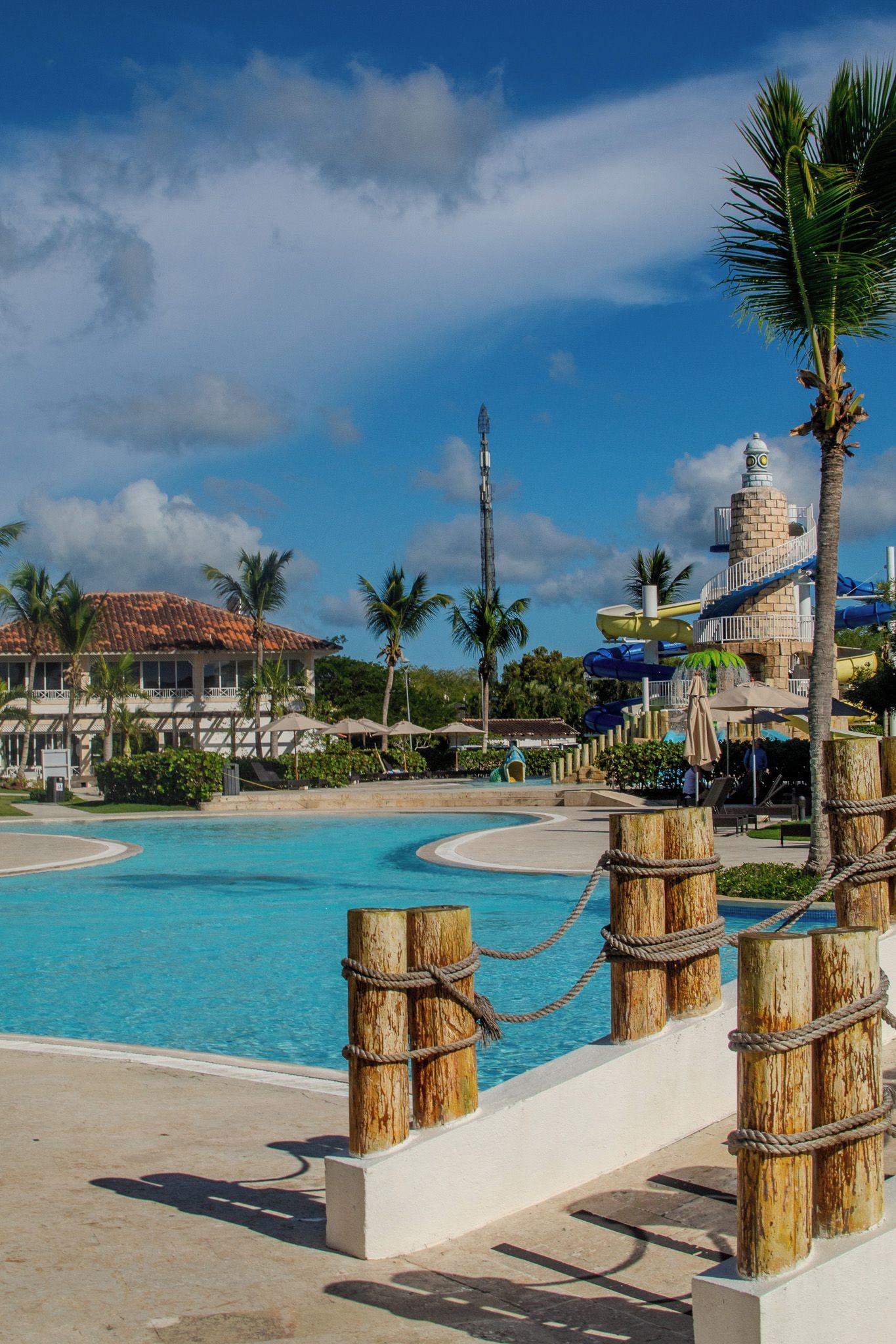 Área de piscina em resort com céu ensolarado, palmeiras e uma estrutura de toboágua.