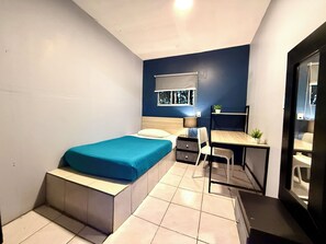 Quarto compacto com cama, mesa de cabeceira, escrivaninha, cadeira e uma pequena janela.