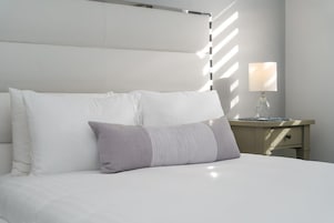 Cama bem arrumada com cabeceira, mesa de cabeceira com abajur, e uma janela na parede projetando sombras.