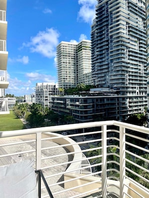 Um edifício residencial alto com várias varandas, céu claro e vista de uma área arborizada com palmeiras.