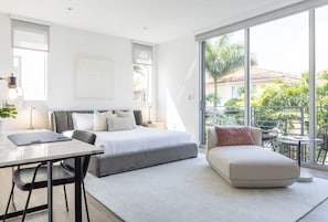 Quarto moderno com cama, sofá, mesa de jantar e uma janela grande com vista para palmeiras.