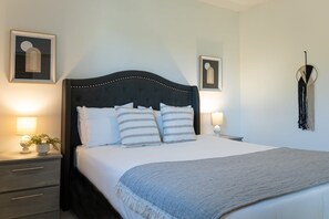Cama bem arrumada com cabeceira escura, duas luminárias de cabeceira e mesa de apoio com luminária e planta.