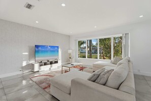 Uma sala de estar moderna com uma televisão de tela plana grande, um sofá, uma mesa de centro e vista para palmeiras através de janelas amplas.