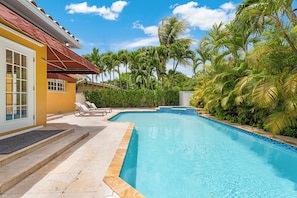 Piscina com borda azulejada, edifício amarelo com toldo vermelho e palmeiras ao fundo.