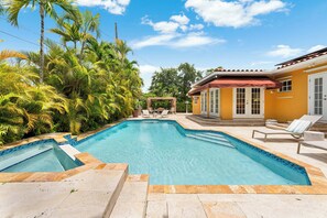 Piscina com borda em azulejo, cercada por palmeiras e um edifício com telhado vermelho.