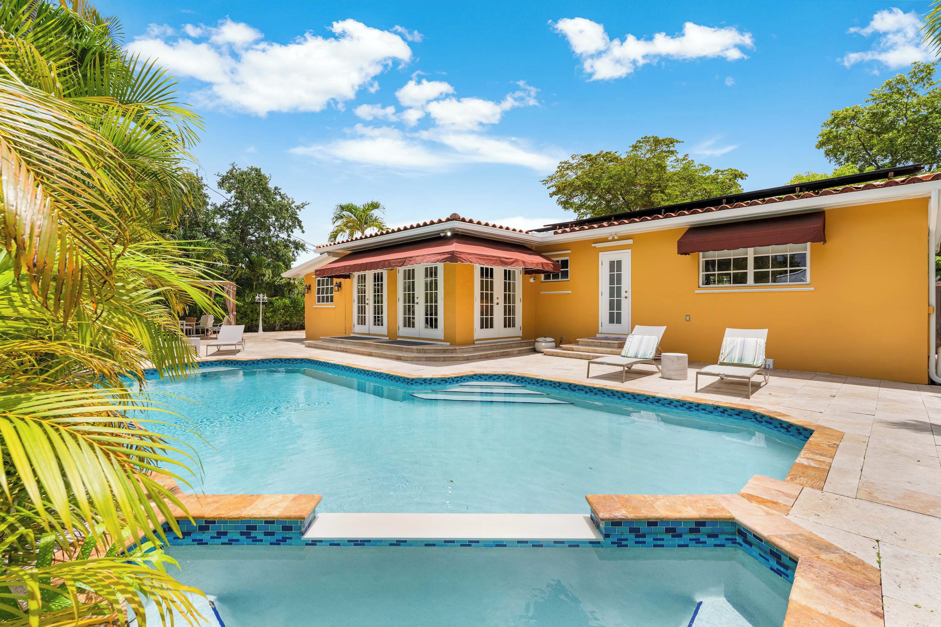 Uma piscina com borda em azulejo, um prédio amarelo com toldo vermelho e uma palmeira.