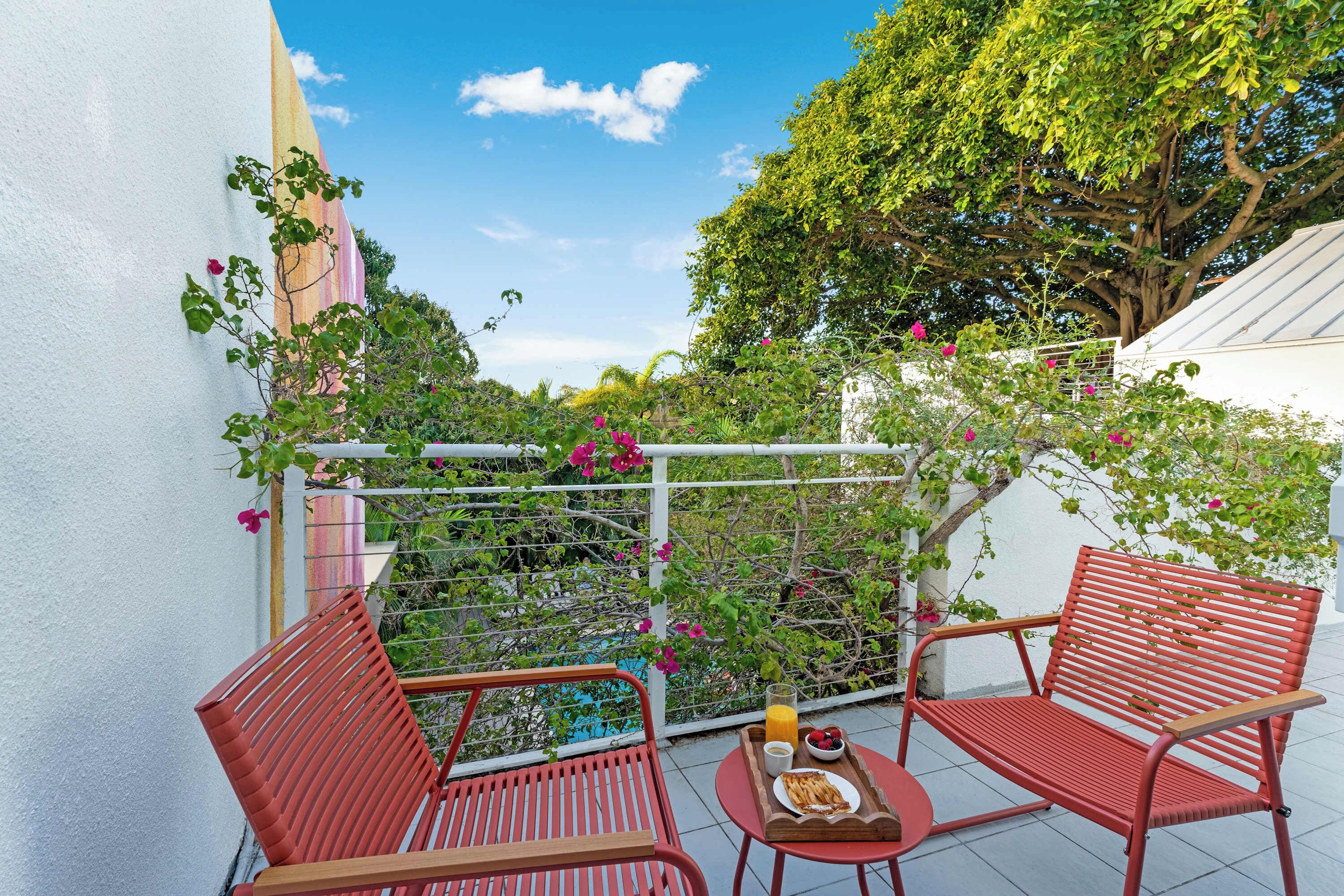 Uma varanda com duas cadeiras de metal vermelhas, uma pequena mesa redonda com uma bandeja de comida e bebidas, e vista para um jardim com árvores e flores.