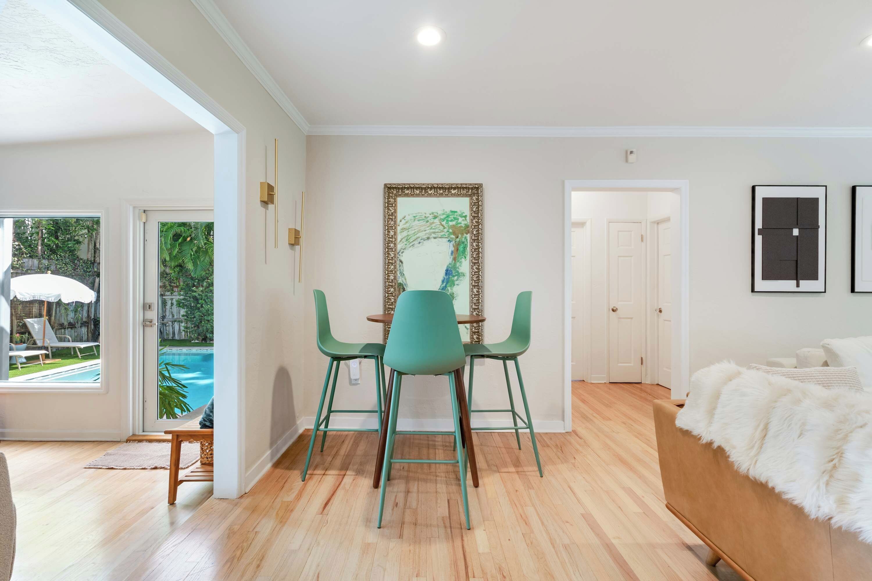 Um quarto com piso de madeira, uma cama com manta branca felpuda, uma mesa de jantar com cadeiras e um quadro na parede.