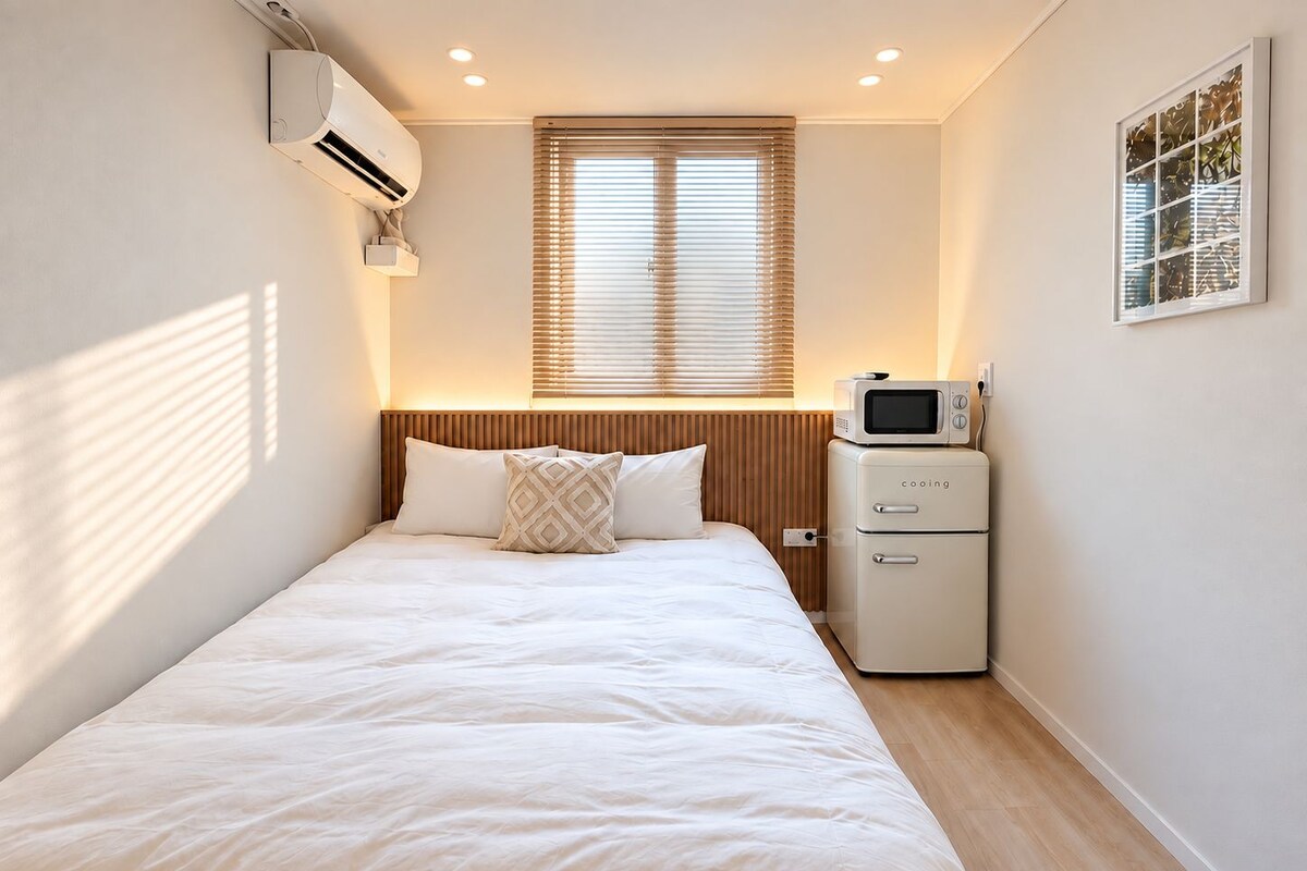 Quarto com cama, mesa de cabeceira, aparelho de ar condicionado, janela com persianas e um quadro na parede.