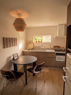 Cozinha compacta com mesa de jantar redonda, duas cadeiras, luminária pendente, janela com plantas e micro-ondas.