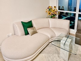 Um sofá seccional bege com uma mesa de centro de vidro, um travesseiro verde e um vaso com flores secas.