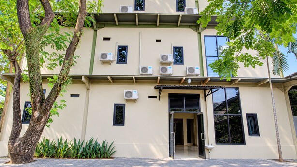 Edifício com várias unidades de ar condicionado, entrada pela porta e área pavimentada com plantas.