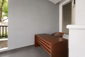 Quarto com escrivaninha de madeira, cadeira, janela com persianas e vista para o verde lá fora.