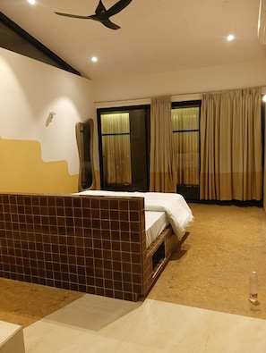 Quarto de hotel com cama, ventilador de teto, janela com cortinas e piso ladrilhado.