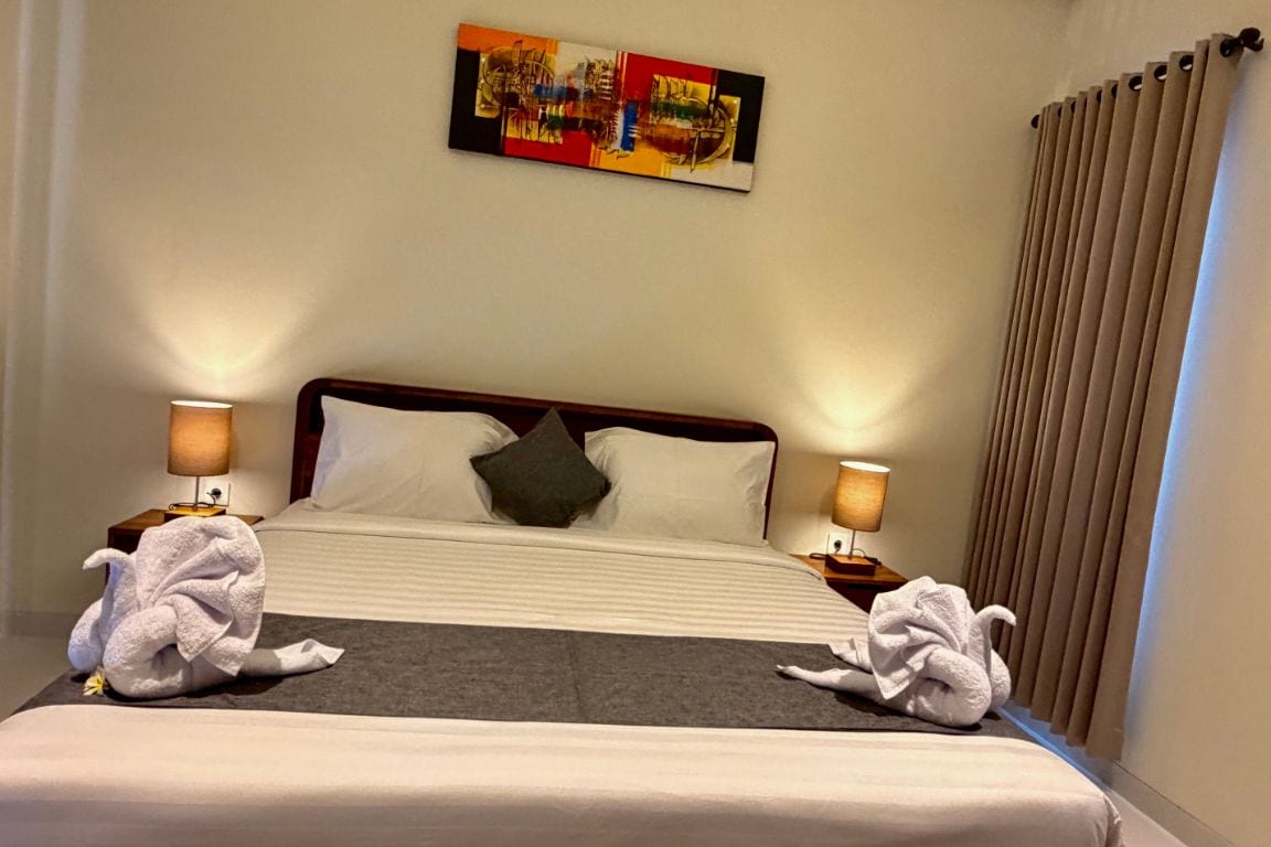 Quarto de hotel com cama, duas luminárias de cabeceira, um quadro na parede e uma janela com cortinas.