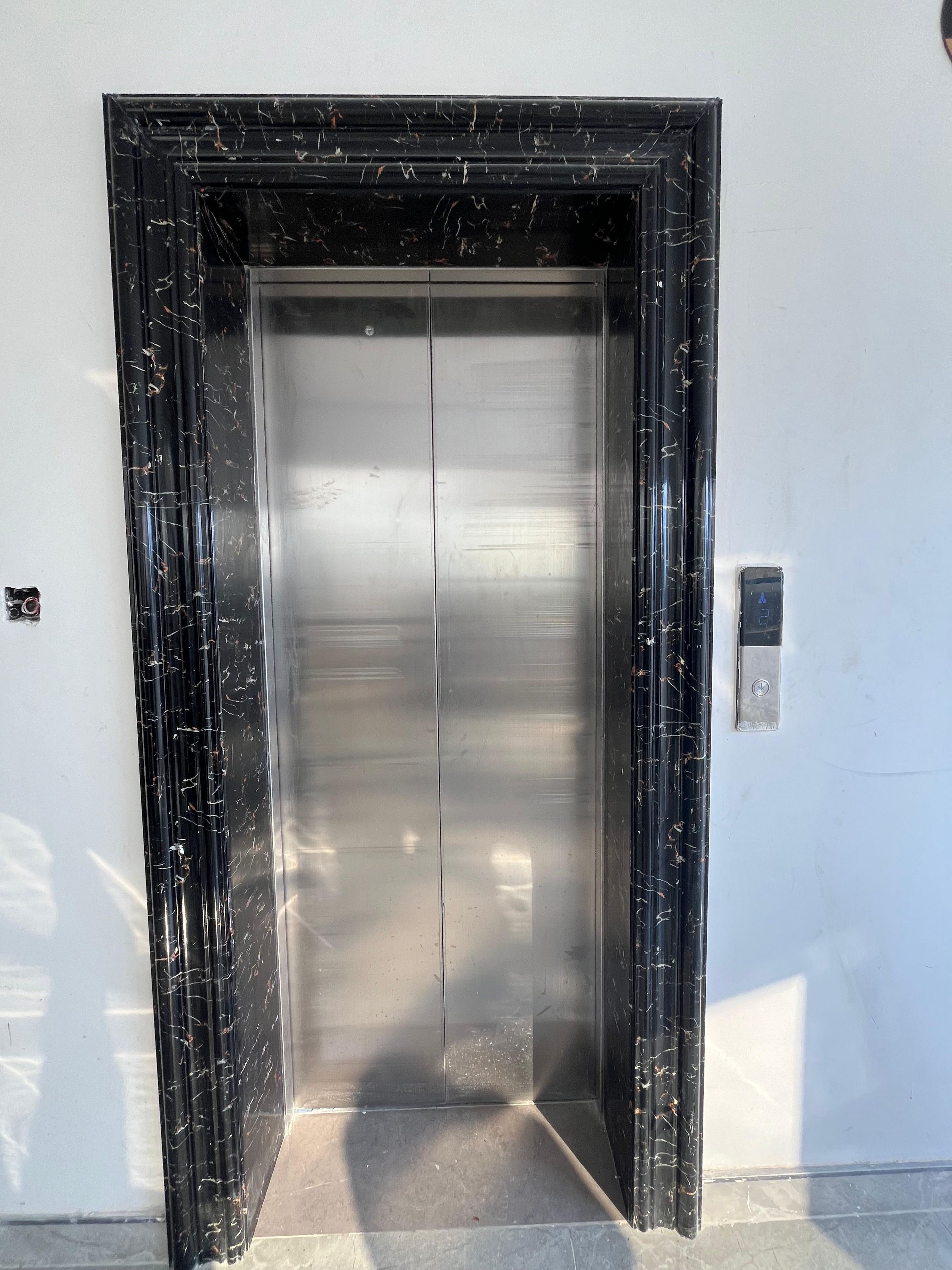 Porta de elevador preta, com interior metálico e painel de controle na parede.