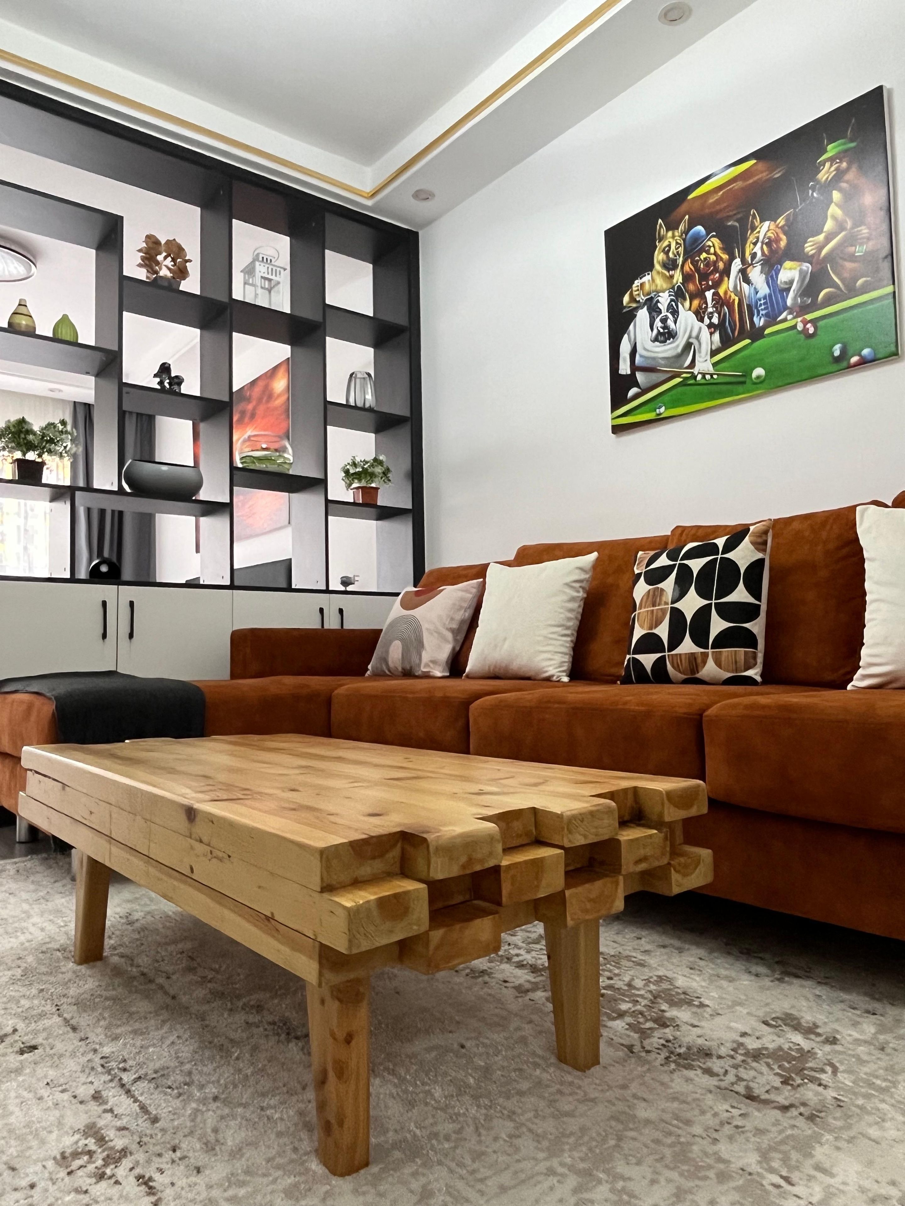 Uma sala de estar moderna com um sofá marrom, uma mesa de centro de madeira, uma estante preta com objetos decorativos e uma pintura de cães jogando sinuca.