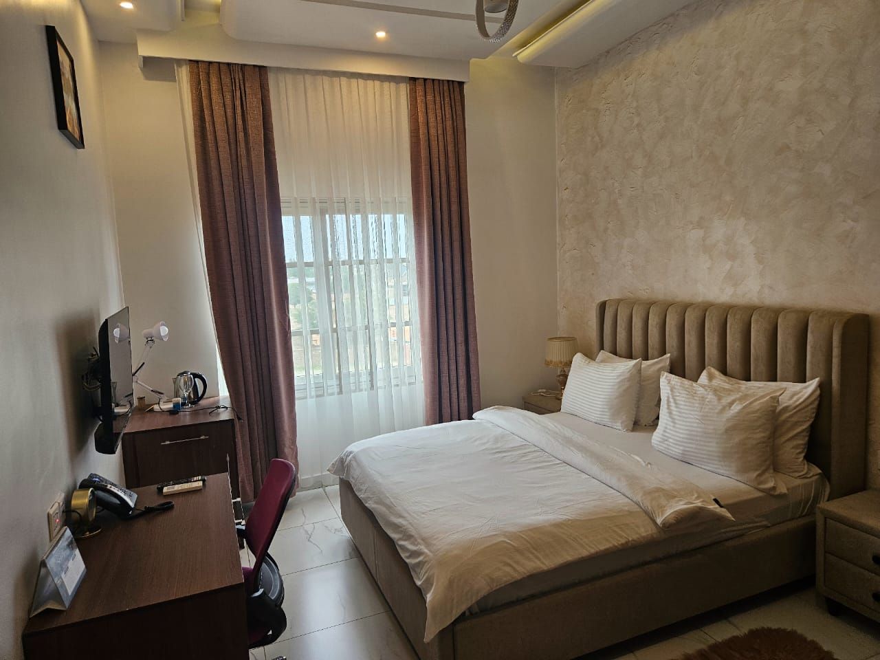 Quarto de hotel com cama, mesa de cabeceira, escrivaninha, cadeira e janela com cortinas.