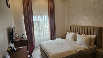 Quarto de hotel com cama, mesa de cabeceira, escrivaninha, cadeira e janela com cortinas.