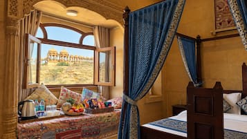 Quarto com cama, janela com vista para paisagem desértica, mesa de cabeceira com cesta de frutas e uma parede decorativa.