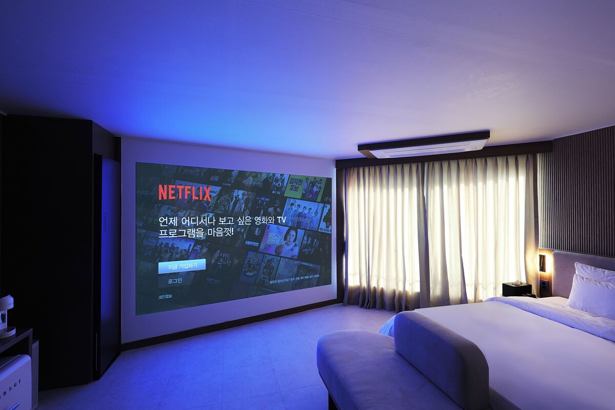 Quarto de hotel com televisão de parede grande exibindo a interface da Netflix, uma cama, uma mesa de cabeceira e uma janela com cortinas.