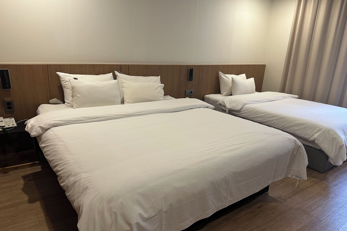 Quarto de hotel com duas camas, cabeceira de madeira, mesa de cabeceira com telefone e cortina.