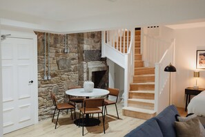 Um quarto aconchegante com parede de pedra, lareira, mesa redonda com cadeiras e escada com corrimãos brancos.