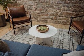 Um quarto com parede de pedra, uma poltrona de couro marrom, uma mesa de centro branca com uma tigela de cobre e um sofá azul.