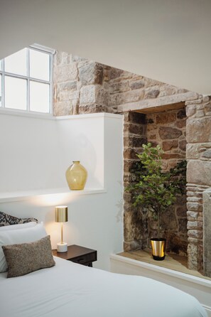 Quarto com parede de pedra, cama, mesa de cabeceira, luminária, vaso e planta.