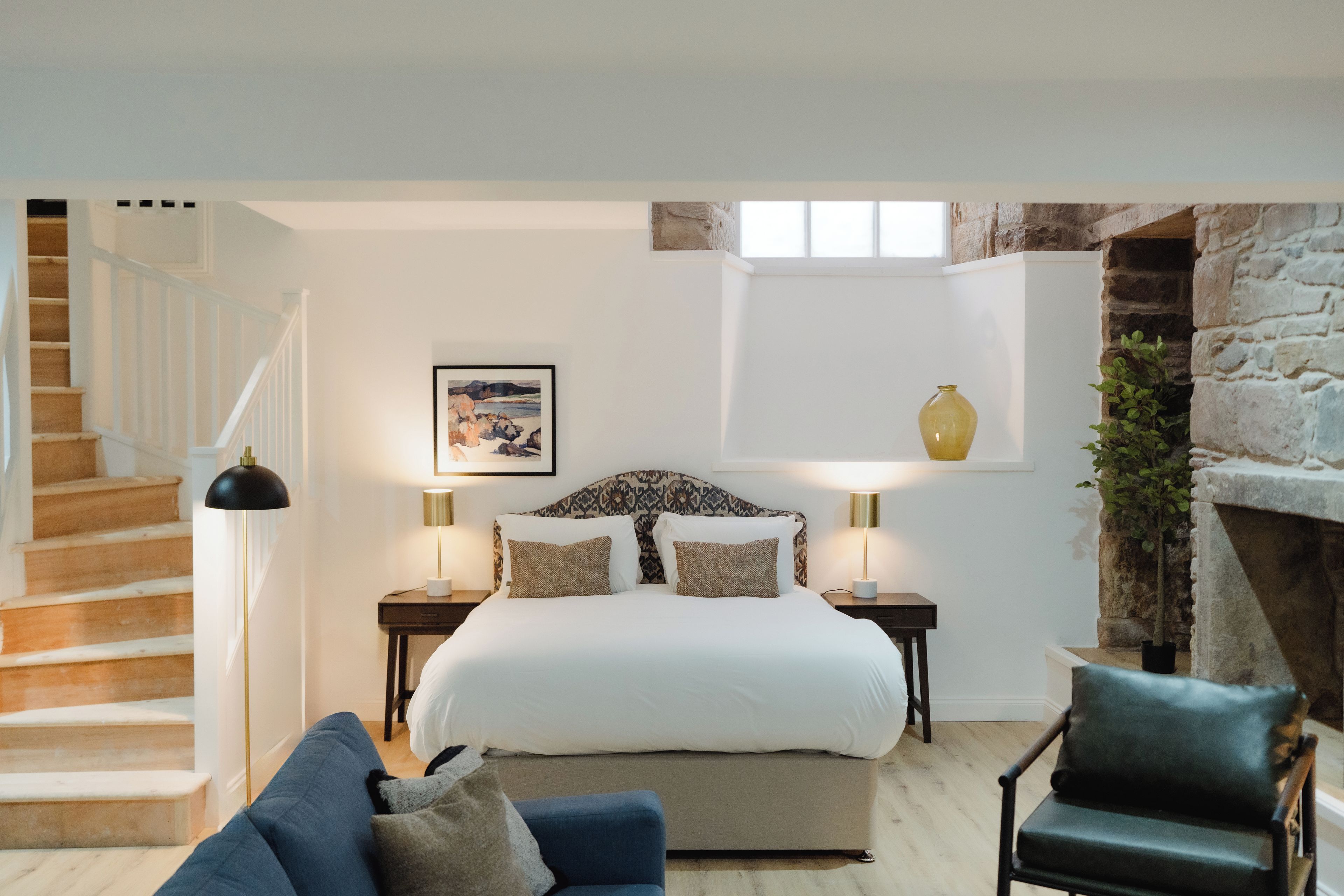 Quarto com cama, duas mesas de cabeceira, luminária, quadro na parede, escada, sofá azul, poltrona e parede de pedra.