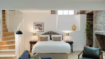 Quarto com cama, duas mesas de cabeceira, luminária, quadro na parede, escada, sofá azul, poltrona e parede de pedra.