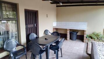 Um pátio externo com mesa e cadeiras, uma lareira de tijolos e um espaço coberto com telhado.