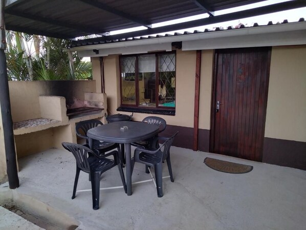 Um pátio externo com uma mesa redonda e cadeiras, uma porta de madeira e uma janela com vista.