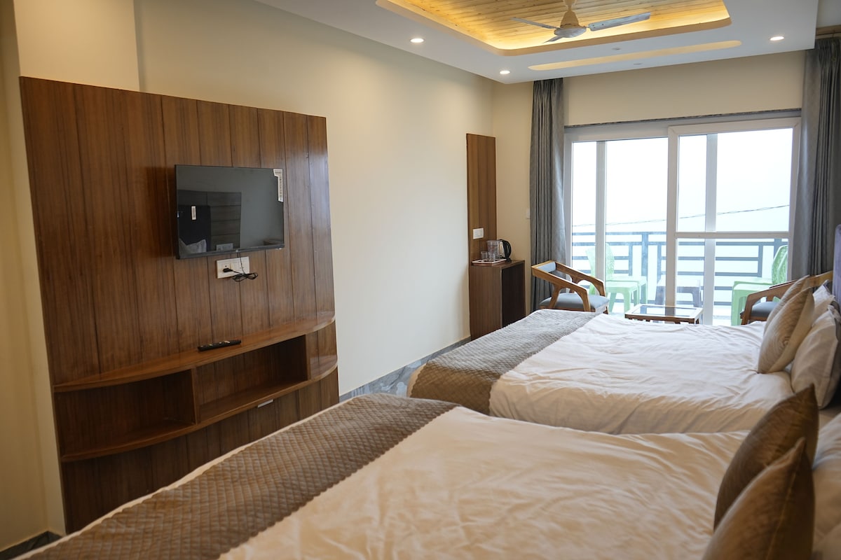 Quarto de hotel com duas camas, uma TV fixada em um painel de madeira, uma mesinha com chaleira e uma varanda com guarda-corpo.