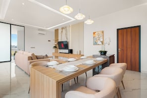 Uma sala de jantar moderna com uma mesa de madeira posta para quatro pessoas, um sofá e uma TV ao fundo.