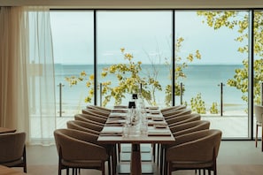 Mesa posta para refeição com cadeiras, janelas amplas com vista para o mar e uma cortina à esquerda.
