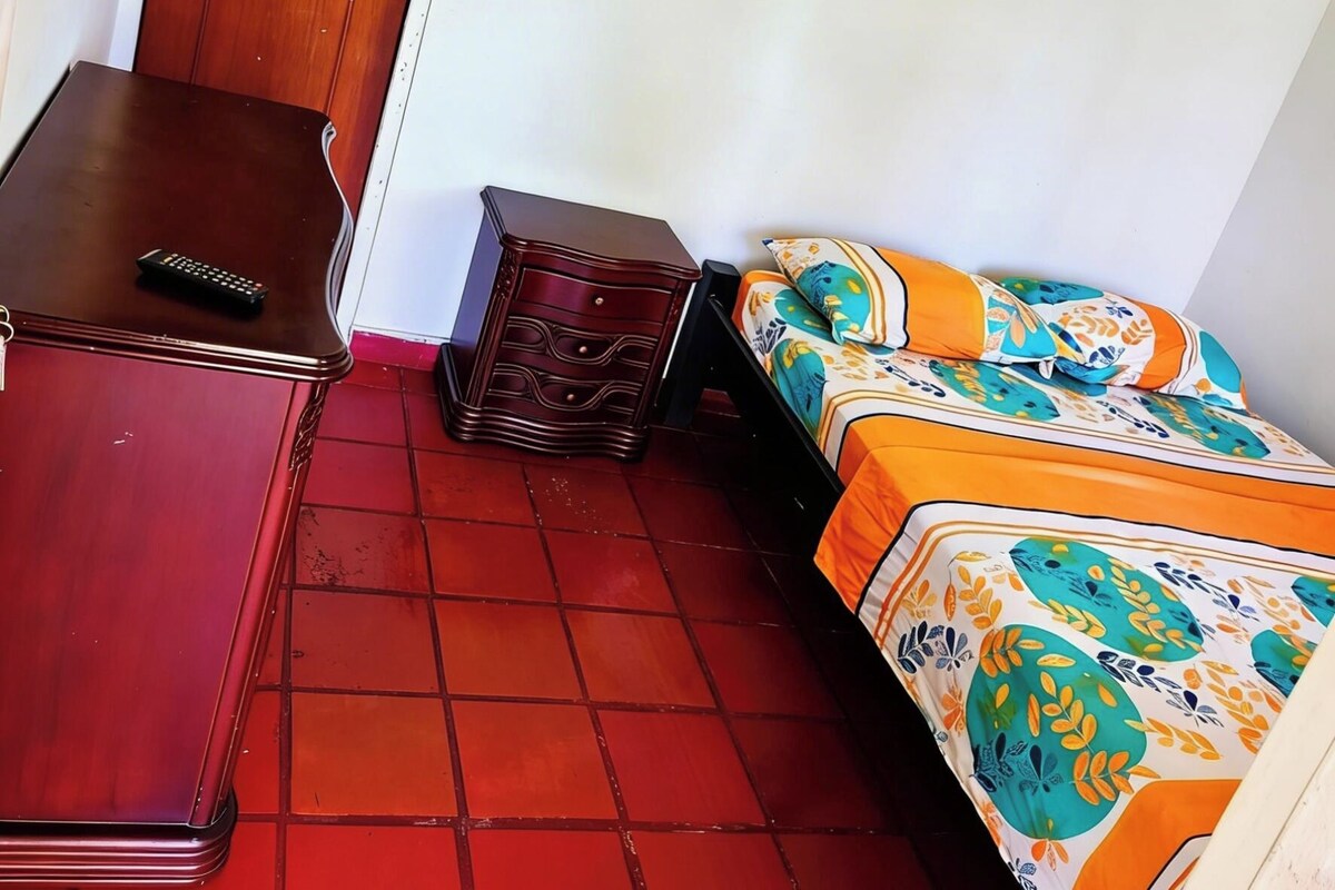 Quarto com porta de madeira, uma mesa pequena com controle remoto, cama com roupa de cama colorida e piso de azulejos.