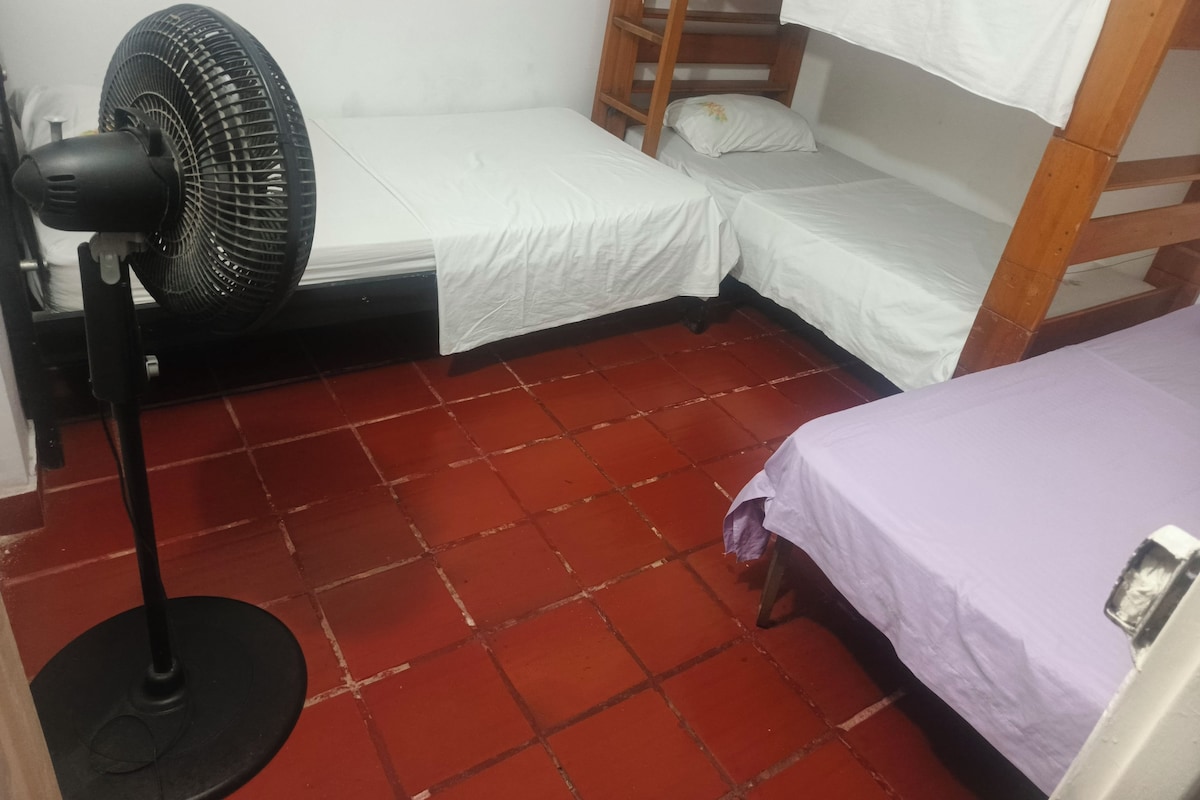 Um quarto pequeno com piso de ladrilho vermelho, uma cama beliche com roupa de cama branca, um ventilador e uma cama de solteiro com roupa de cama roxa.