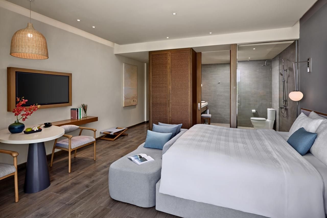 Quarto de hotel moderno com uma cama grande, área de estar com mesa e cadeiras, televisão e banheiro com chuveiro e vaso sanitário.