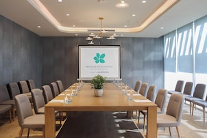 Uma sala de conferências com uma mesa comprida, cadeiras, uma tela de projetor e um vaso de plantas.