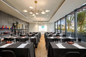 Sala de conferências com mesas preparadas para reuniões, uma tela de projetor e vista para palmeiras através de grandes janelas.