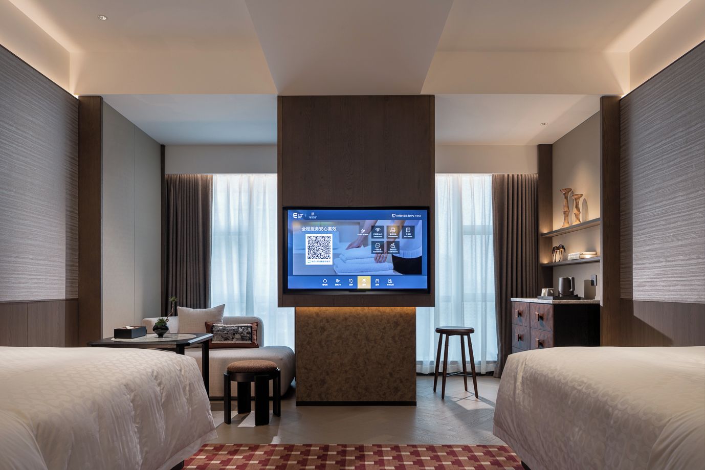Quarto de hotel com duas camas, televisão fixada na parede, mesa pequena com uma cadeira, sofá e armário com objetos decorativos.