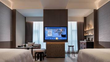 Quarto de hotel com duas camas, televisão fixada na parede, mesa pequena com uma cadeira, sofá e armário com objetos decorativos.