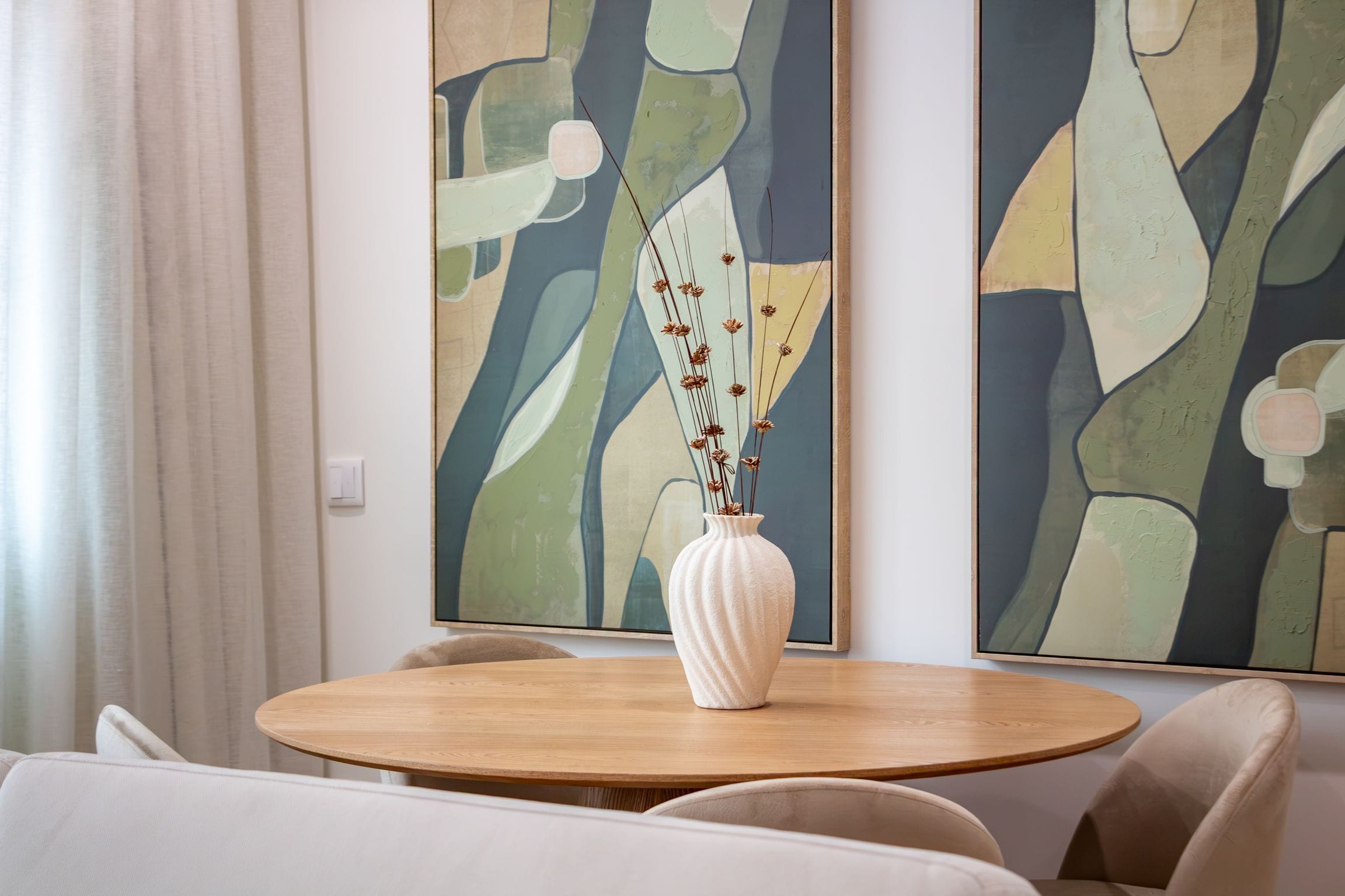 Quarto com uma mesa de madeira redonda, um vaso branco com flores secas e quadros abstratos na parede.