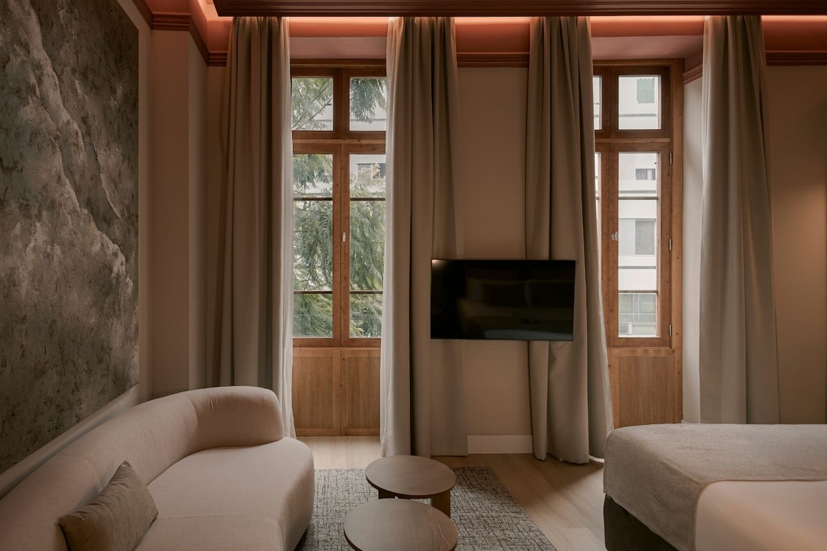 Quarto de hotel com uma grande parede de pedra, um sofá, uma cama, uma TV e janelas amplas com cortinas.
