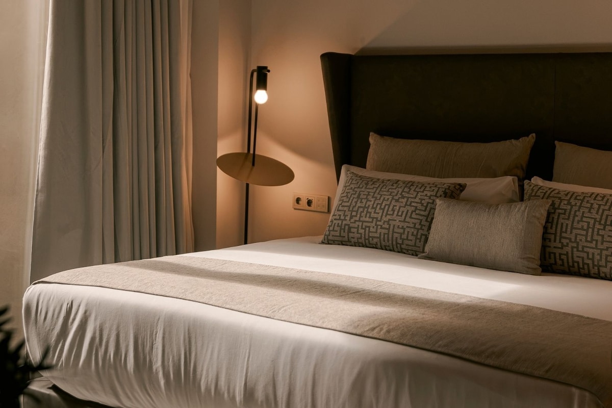 Quarto de hotel com uma cama grande, abajur e cabeceira com várias almofadas.