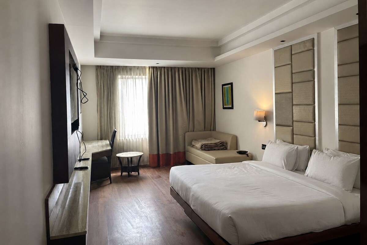 Quarto de hotel com cama, escrivaninha, cadeira, mesa pequena e televisão.