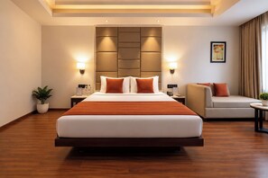 Quarto de hotel moderno com uma cama grande, um sofá, um painel de luz fixado na parede, um vaso de planta e um quadro na parede.