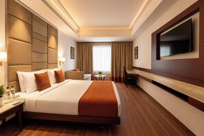 Quarto de hotel com uma cama grande, uma escrivaninha, uma cadeira, uma televisão e uma janela com cortinas.