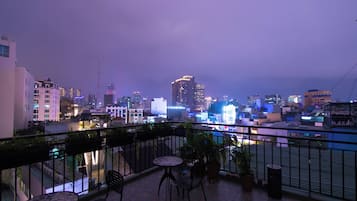Varanda com cadeiras e mesa, vista para um horizonte urbano com edifícios iluminados ao anoitecer.